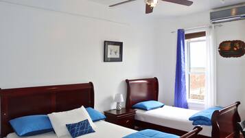 Quarto de hotel com duas camas de solteiro, um ventilador de teto, uma janela com cortinas e uma placa na parede.