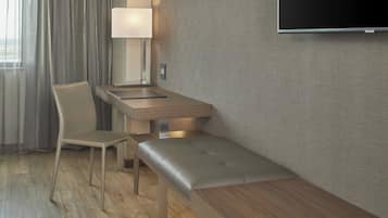 Quarto de hotel moderno com escrivaninha de madeira, um banco acolchoado, uma cadeira, abajur e TV de tela plana fixada na parede.