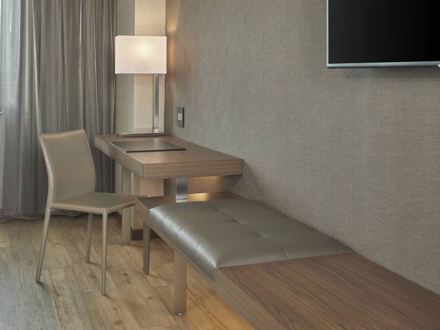 Quarto de hotel moderno com escrivaninha de madeira, um banco acolchoado, uma cadeira, abajur e TV de tela plana fixada na parede.