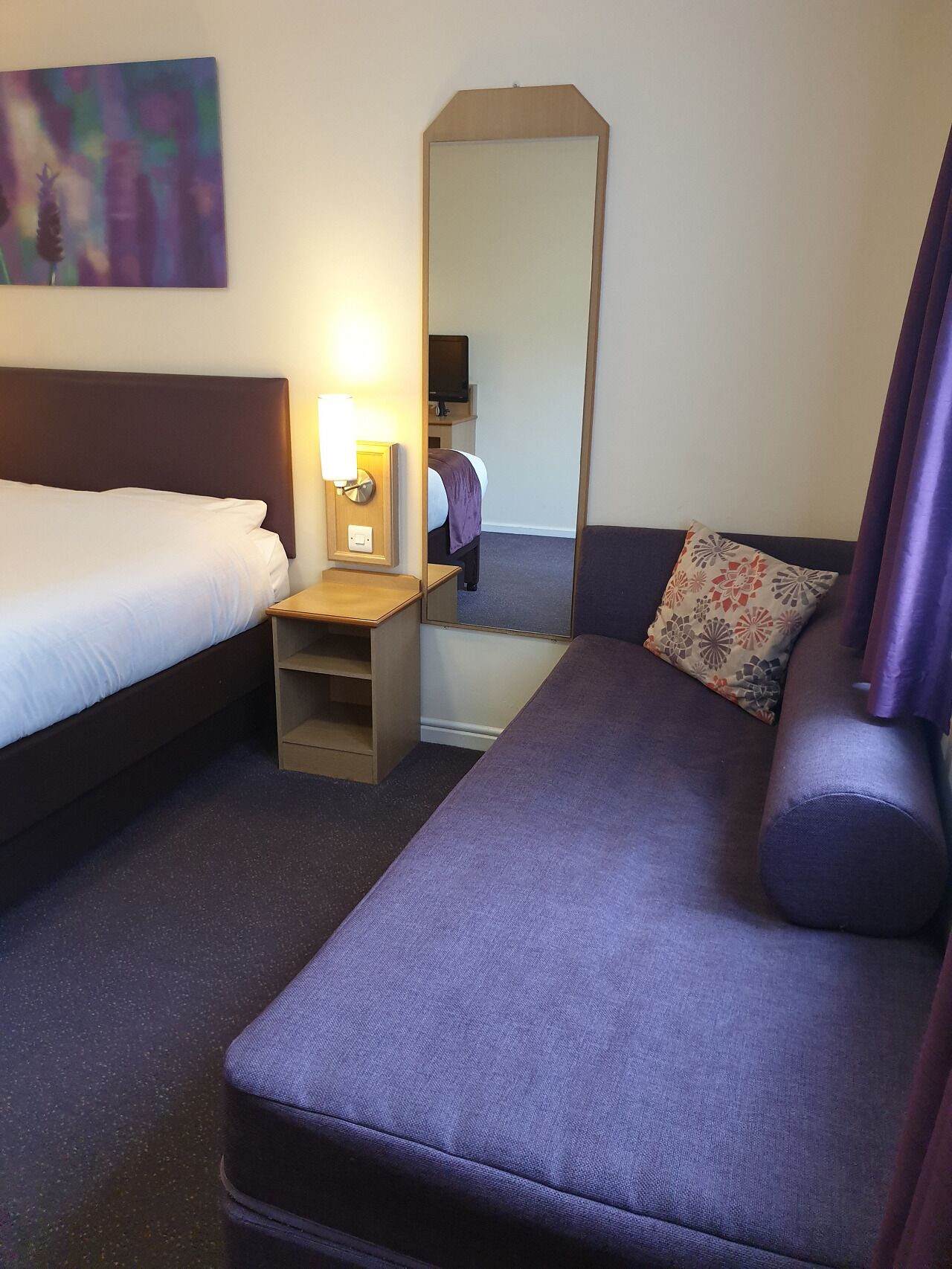 Quarto de hotel com cama, sofá, mesa de cabeceira com abajur e um espelho grande.