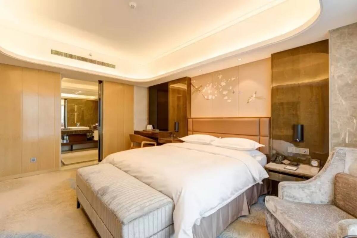 Quarto de hotel moderno com uma cama grande, área de estar e banheiro visível por uma porta aberta.