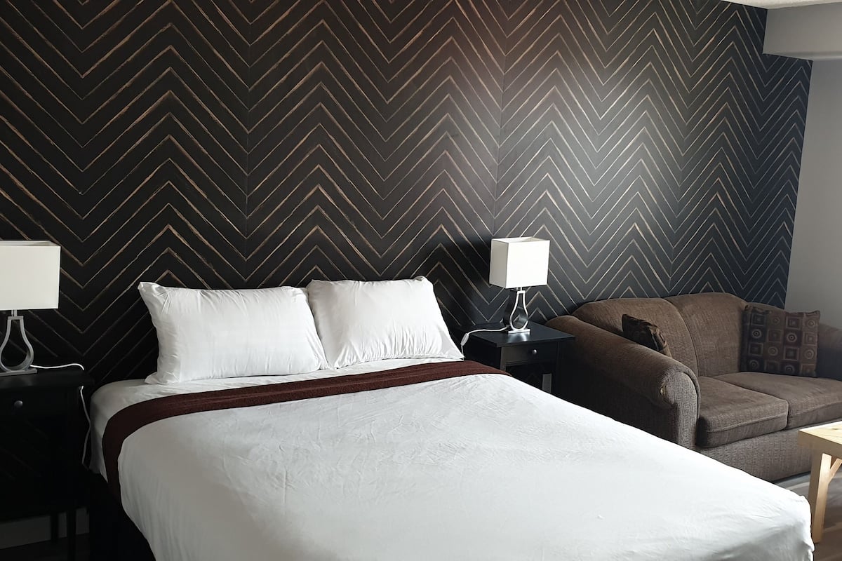 Quarto de hotel com uma cama, duas mesas de cabeceira com luminárias, um sofá, uma pequena mesa de madeira e uma parede estampada.
