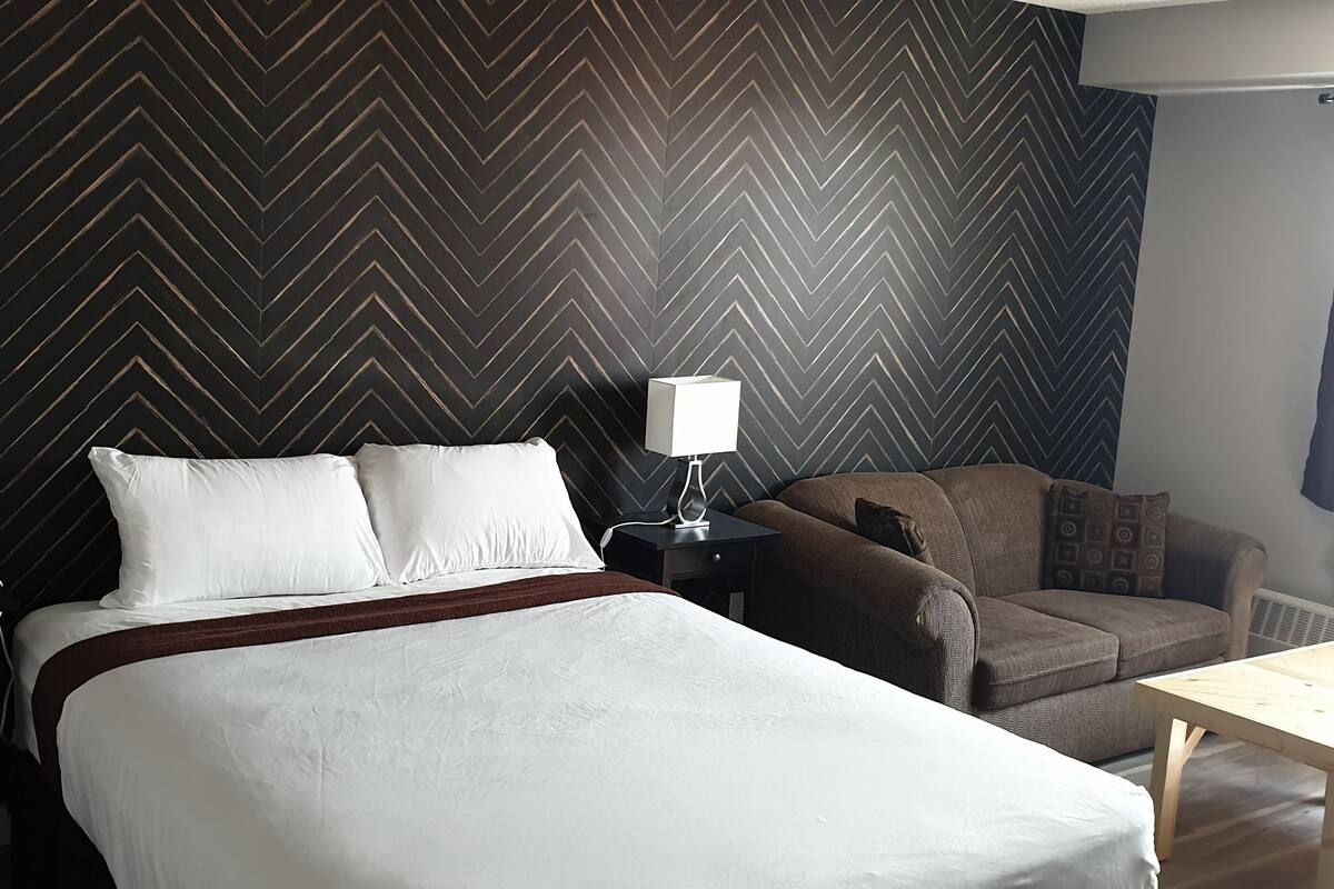 Quarto de hotel com cama, sofá, mesa de cabeceira com luminária, mesa e parede com padrão em zigue-zague.