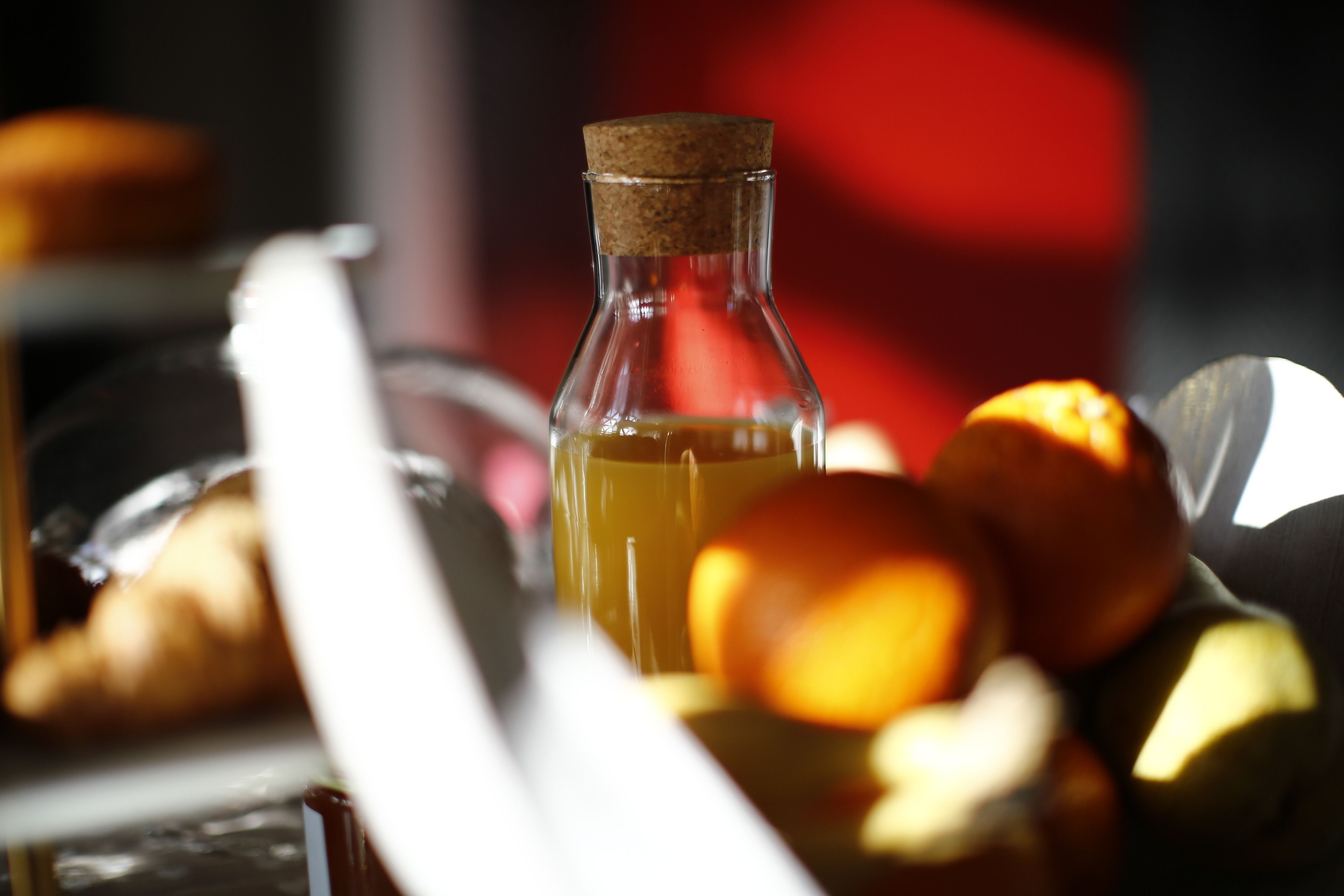 Uma garrafa de vidro com rolha, um copo de suco de laranja e uma tigela com frutas, incluindo laranjas e maçãs.
