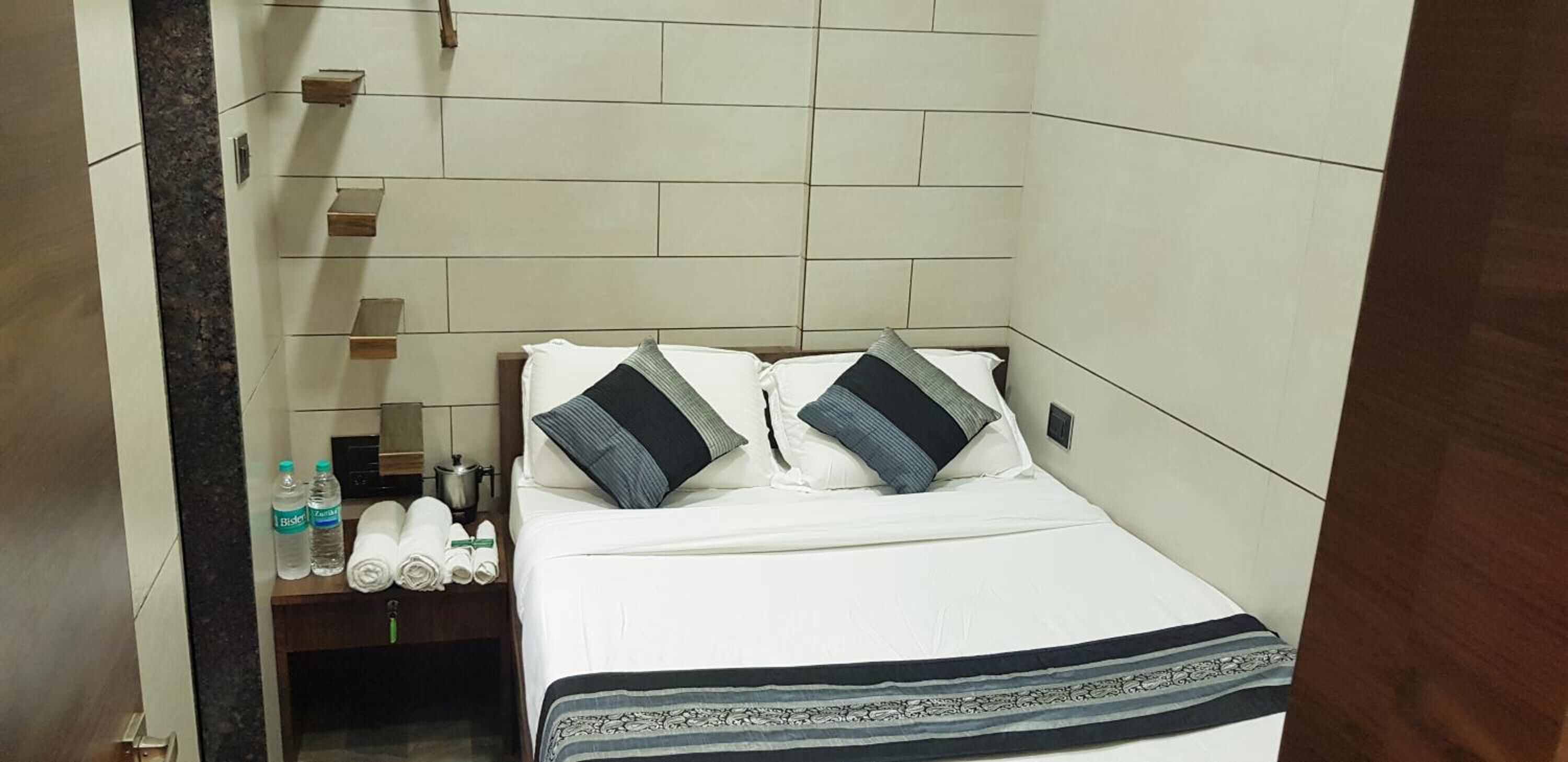 Quarto de hotel com cama, almofadas, mesa de cabeceira com artigos de higiene e parede revestida de azulejos.