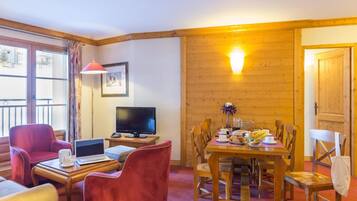 Um quarto aconchegante com parede de madeira, uma mesa de jantar posta com cadeiras, uma televisão e um laptop sobre uma mesinha.