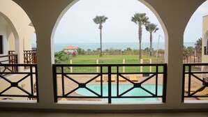 Vista através de uma varanda com piscina, palmeiras e céu claro.
