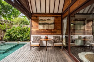 Um deck de madeira com piscina, duas cadeiras, uma mesa com frutas e um painel de parede tecido.