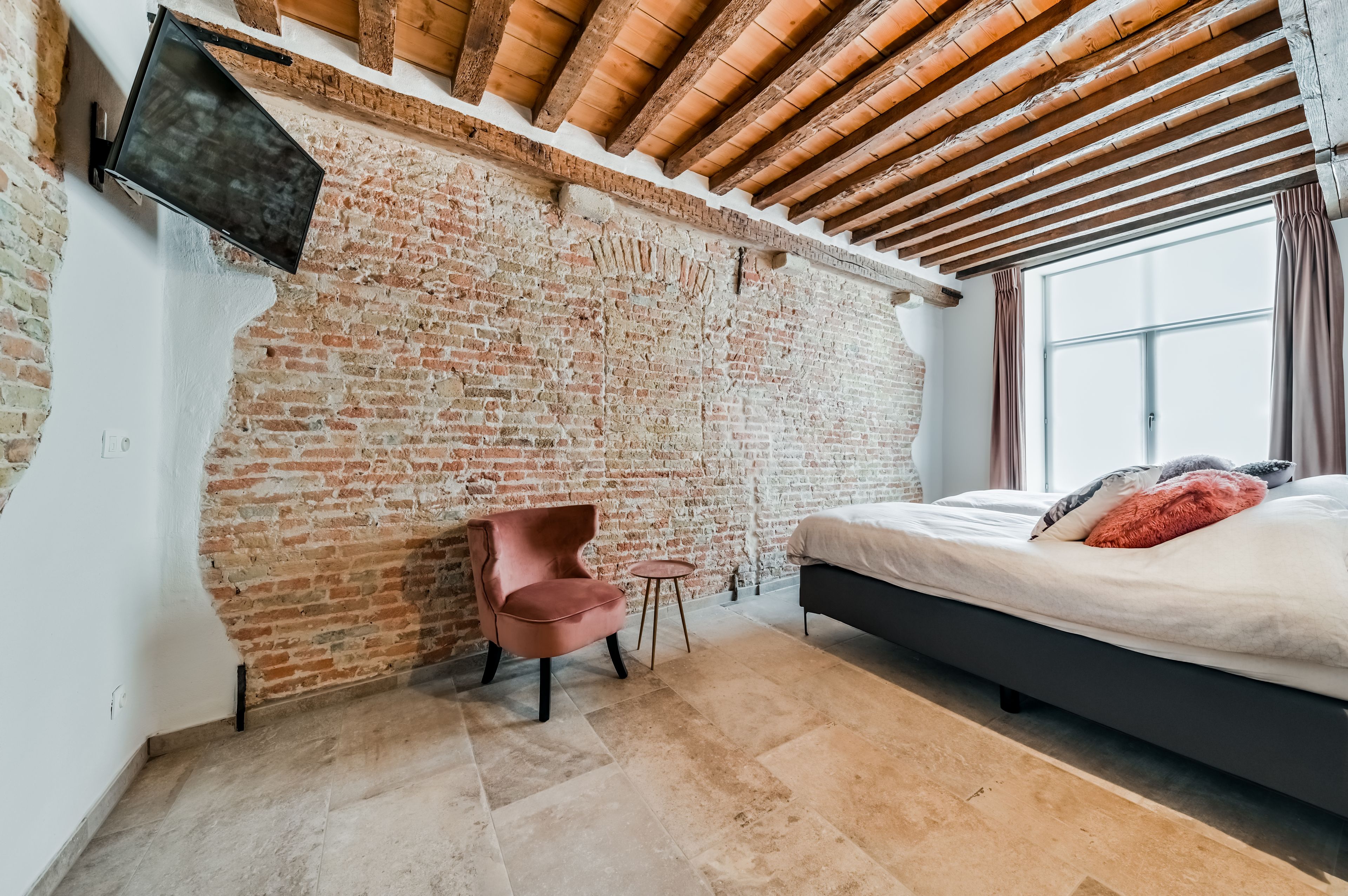 Quarto com paredes de tijolo aparente, teto de madeira, uma cama, uma cadeira e uma pequena mesa.