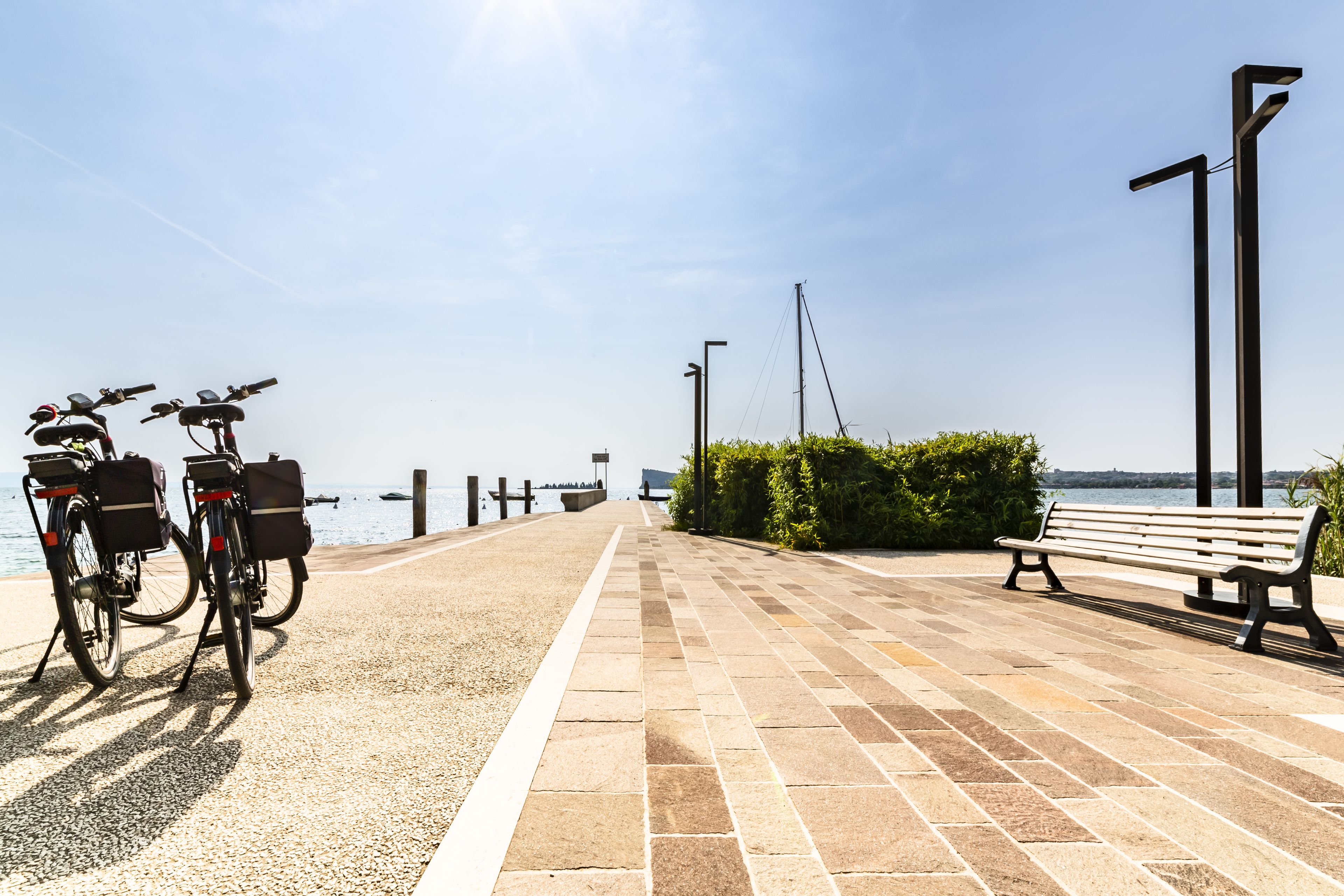 Uma passarela pavimentada, bicicletas estacionadas à esquerda, um banco à direita e vista da água ao longe.