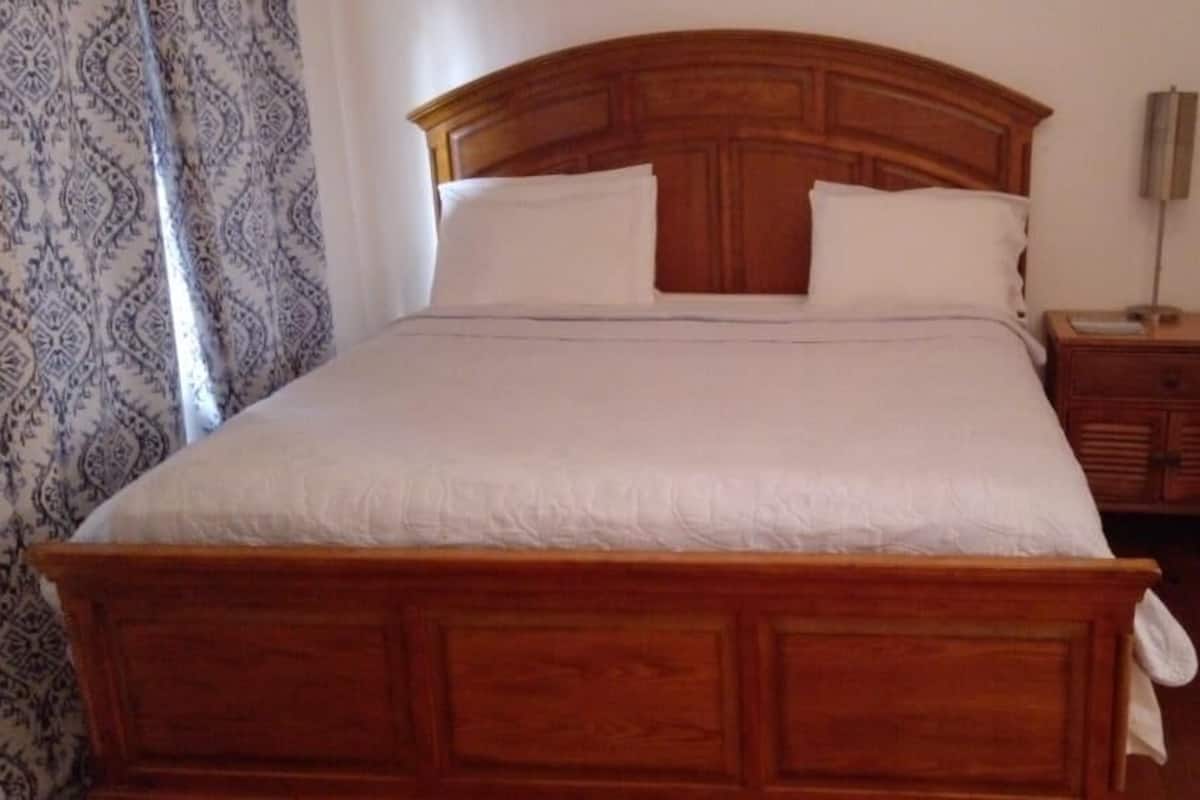 Uma cama de madeira com cabeceira, duas almofadas, mesa de cabeceira e uma luminária.