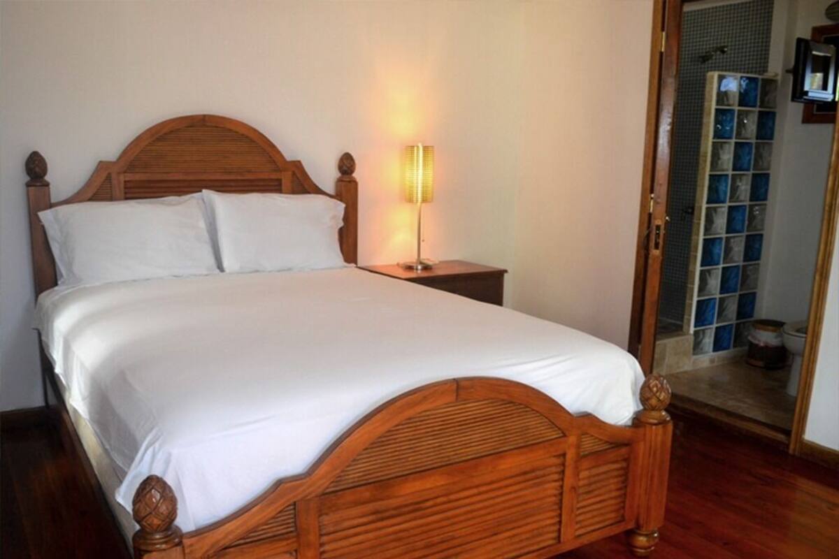 Uma cama bem arrumada com cabeceira de madeira, abajur de cabeceira e vista para um banheiro com box revestido em azulejo.