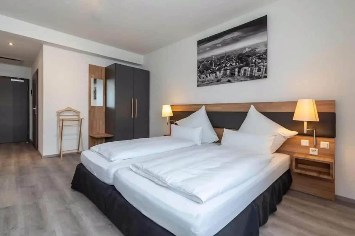 Uma cama de casal com roupa de cama branca, duas mesas de cabeceira com abajures, uma cabeceira, um guarda-roupa e um quadro na parede.