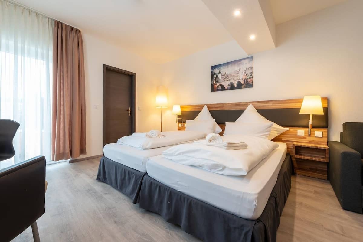 Quarto de hotel com uma cama de casal, duas mesas de cabeceira com abajures, uma cadeira, uma escrivaninha e uma porta.