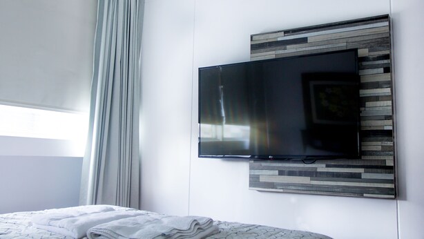 Quarto de hotel moderno com televisão de tela plana fixada em parede com design em madeira e metal, cama bem arrumada com colcha estampada e uma janela grande com cortinas.