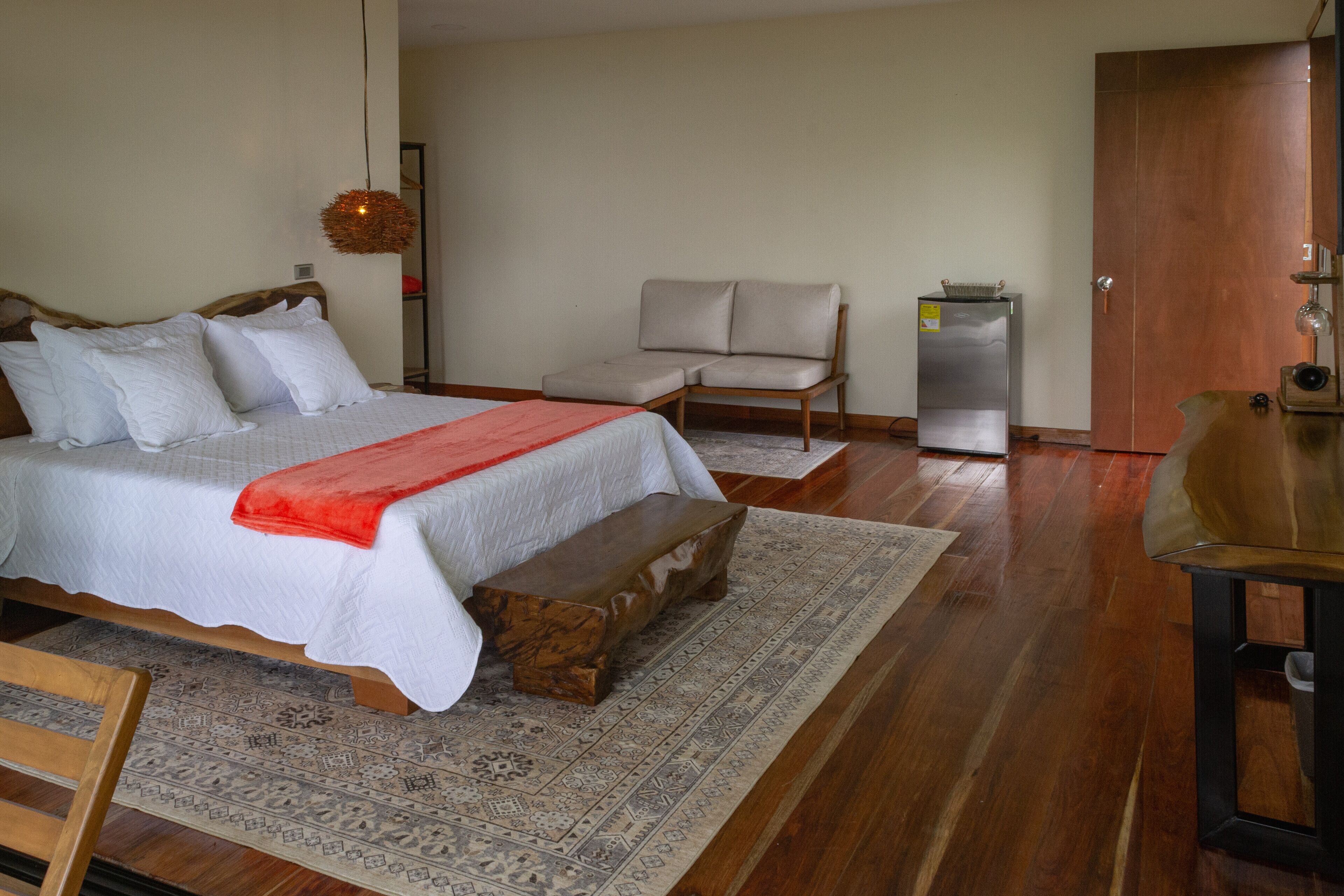 Quarto com cama, um banquinho, uma cadeira, geladeira e porta de madeira.