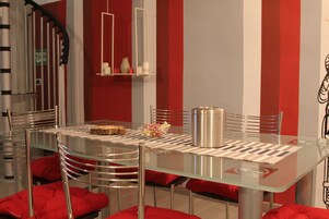 Uma área de refeições moderna com mesa de vidro, cadeiras de metal e almofadas vermelhas.