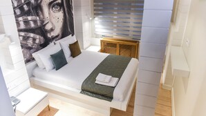 Quarto moderno com uma cama grande, uma mesinha de cabeceira, um pequeno baú de madeira e uma obra de arte na parede.