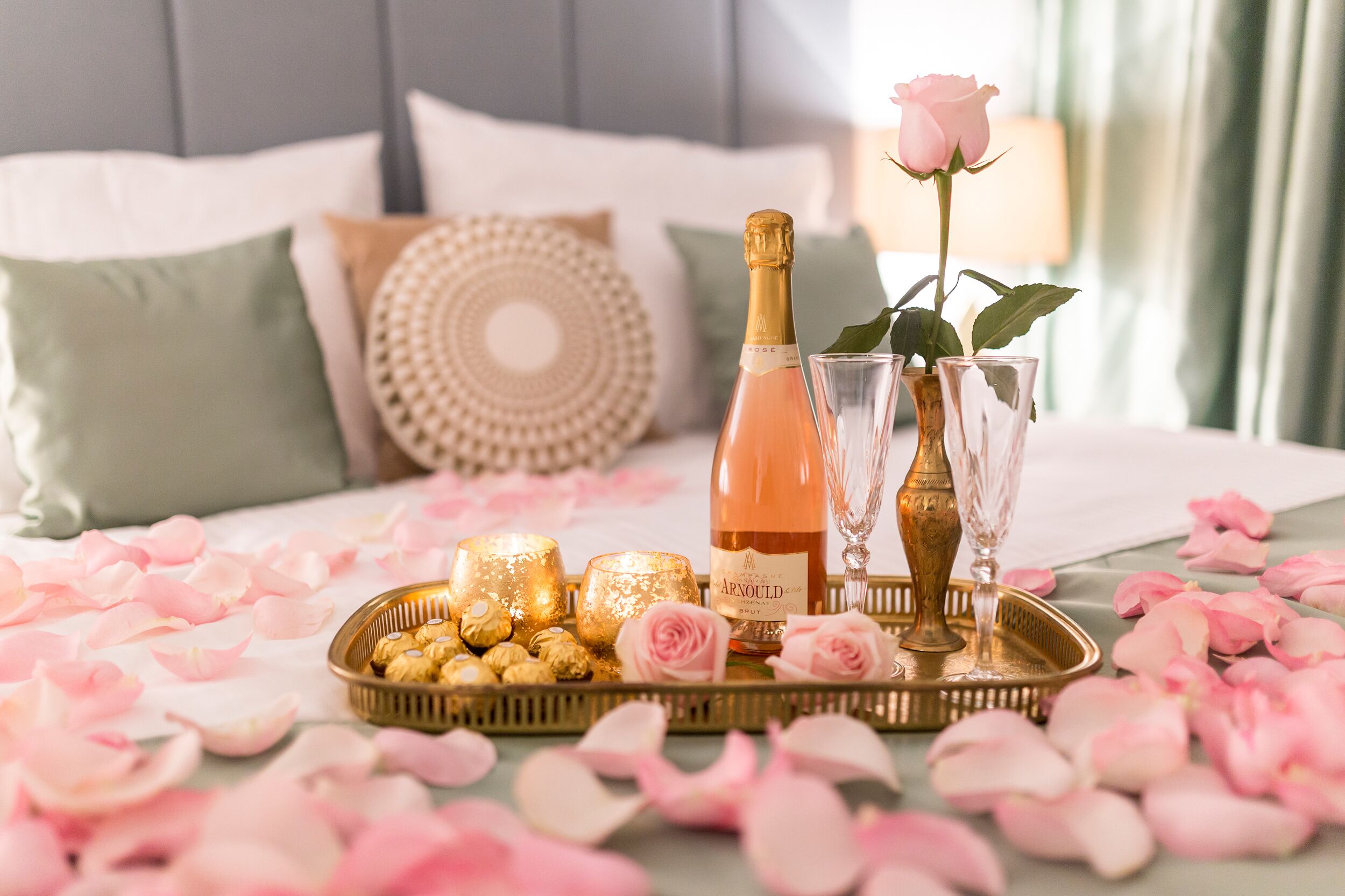 Uma cama com uma garrafa de champanhe, duas taças, um tabuleiro com chocolates e pétalas de rosas.