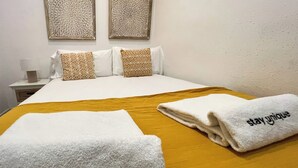 Cama bem arrumada com colcha cor mostarda e duas almofadas, uma com desenho estampado. Há duas toalhas na cama, uma com a frase 'stay unique' e outra com o mesmo texto.