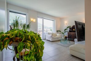 Uma sala de estar moderna com sofá branco, mesa de centro de vidro e TV. Há janelas amplas com vista para uma montanha e uma varanda.