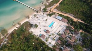 Vista aérea de um resort na praia, com um longo píer, uma piscina e vários edifícios no estilo de bangalô.