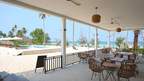 Área de estar coberta ao ar livre com mesas e cadeiras, com vista para uma praia e piscina.