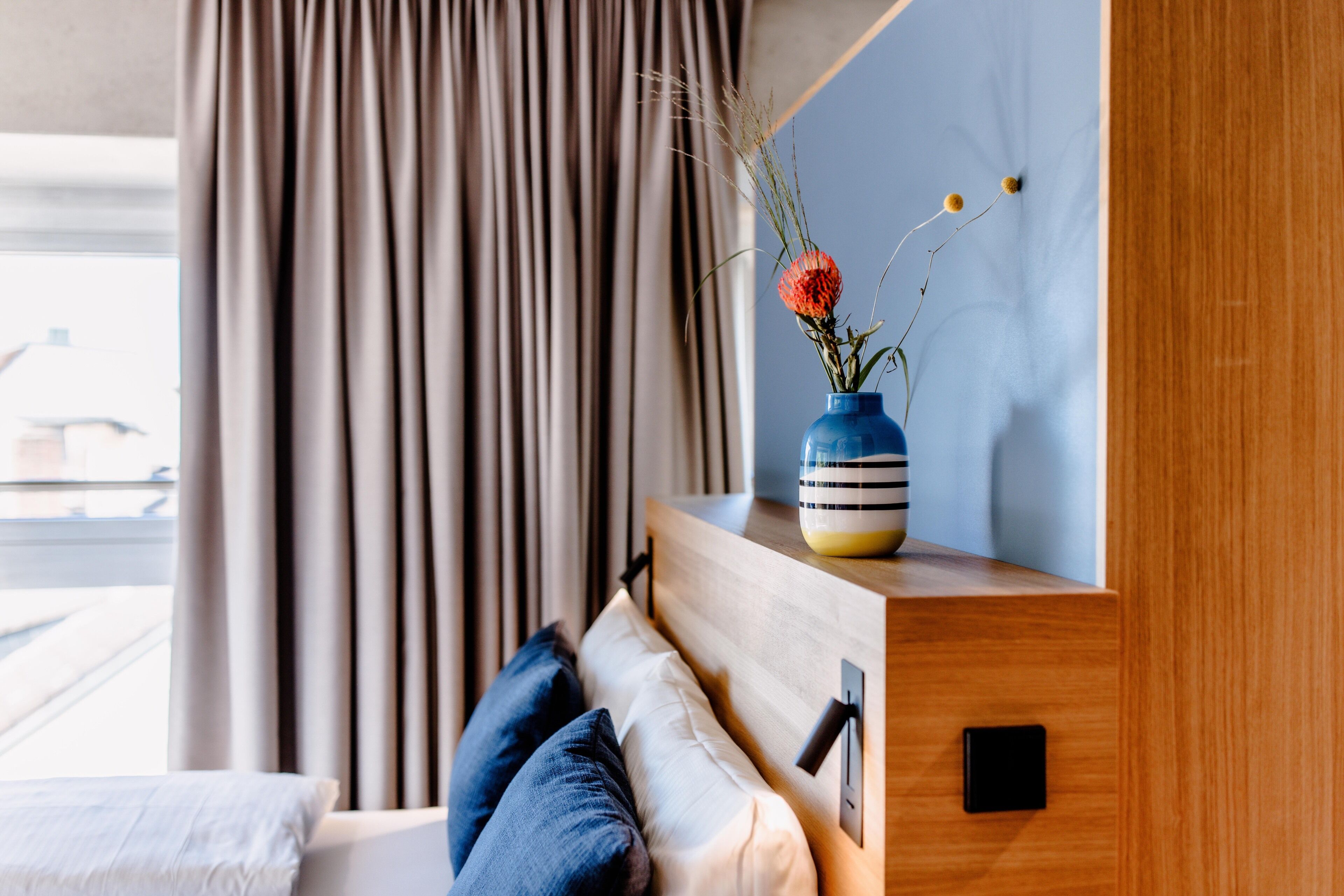 Quarto moderno com cabeceira de madeira, cama com roupa de cama azul e branca, um vaso com flores em uma mesa de cabeceira e vista para o exterior através de grandes janelas.