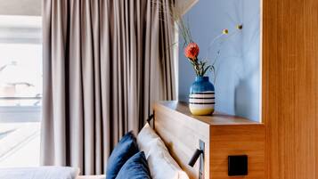 Quarto moderno com cabeceira de madeira, cama com roupa de cama azul e branca, um vaso com flores em uma mesa de cabeceira e vista para o exterior através de grandes janelas.