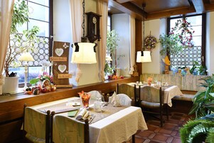 Uma área de refeições aconchegante com móveis de madeira, toalhas de mesa brancas e diversas plantas.