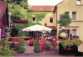 Um café ao ar livre com mesas e cadeiras, rodeado por vegetação e um edifício com uma placa.