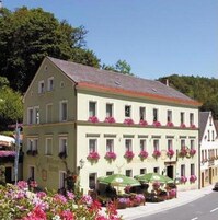 Um edifício de dois andares com telhado plano e várias janelas, algumas com floreiras. Há mesas ao ar livre com guarda-sóis.