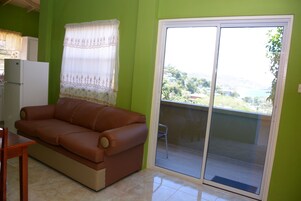 Quarto com sofá de couro marrom, geladeira branca e porta de vidro corrediça que dá para uma varanda com vista para vegetação e edifícios.