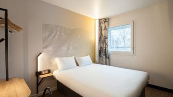 Quarto de hotel com uma cama grande, uma mesa de cabeceira com abajur, um cabideiro e uma janela com cortinas.