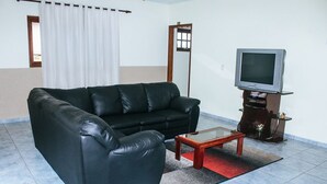 Sala de estar com sofá de couro preto, mesa de centro de vidro, televisão sobre suporte de madeira e tapete vermelho.