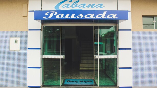 Entrada de um hotel chamado "Cabana Pousada", com porta de vidro e uma placa azul e branca acima.