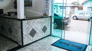 Uma área de recepção com balcão de mármore, porta de vidro que dá para o exterior e tapete azul com a inscrição 'Bem Vindo'.