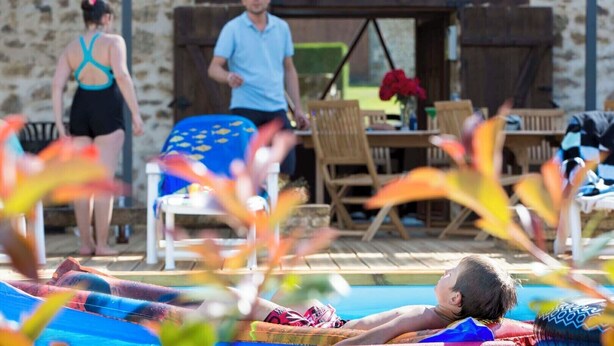Uma área ao lado da piscina com uma mesa de jantar e cadeiras, uma pessoa caminhando e outra relaxando em uma espreguiçadeira.
