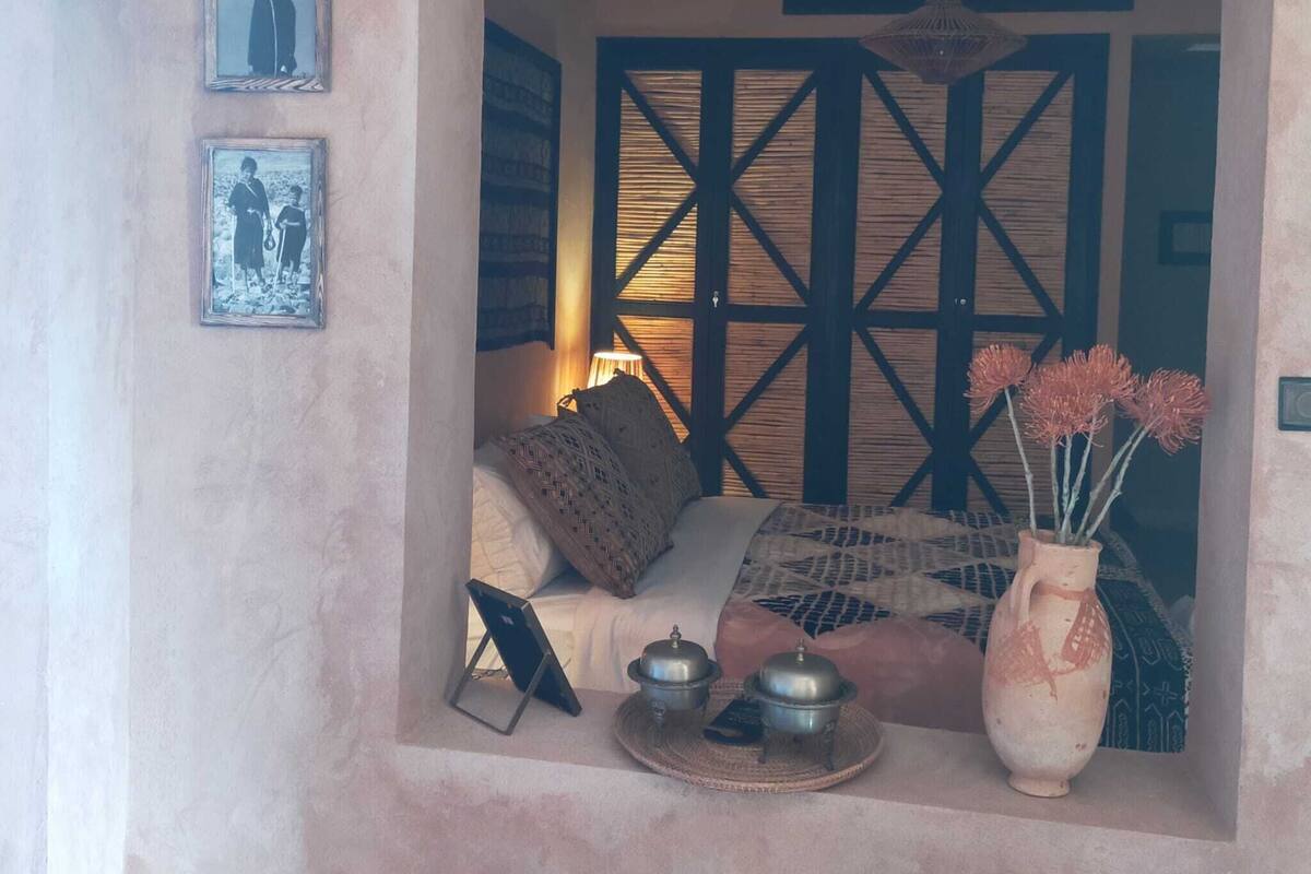 Quarto aconchegante com cama, almofadas, uma mesinha com um vaso e quadros nas paredes.