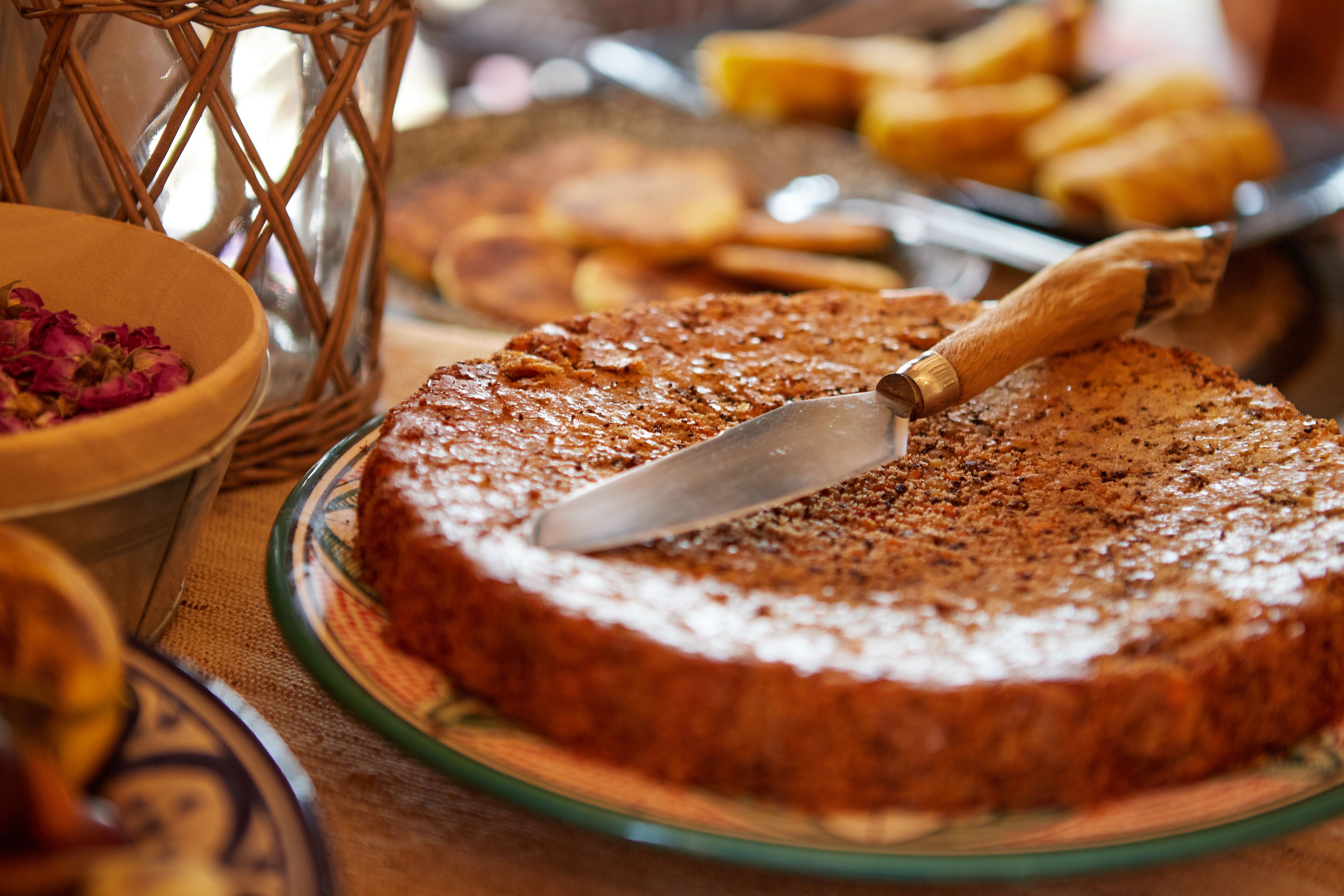 Bolo redondo com cabo de madeira sobre um prato de vidro, rodeado por outros alimentos e um frasco de vidro decorativo.