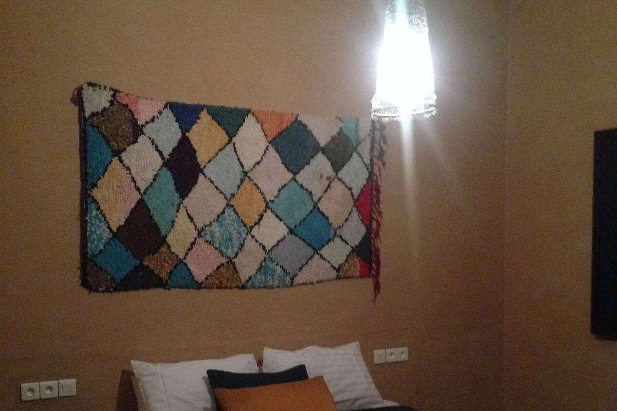 Quarto com cama, almofadas, colcha colorida e luminária na parede.