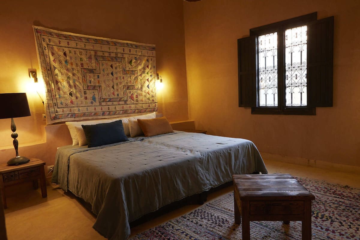 Um quarto com uma cama grande, duas luminárias laterais, um tapeçaria na parede e uma janela com grades de ferro.