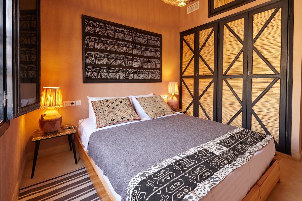 Uma cama com almofadas estampadas, uma obra de arte na parede, uma mesa de cabeceira com abajur e uma janela com persianas de madeira.