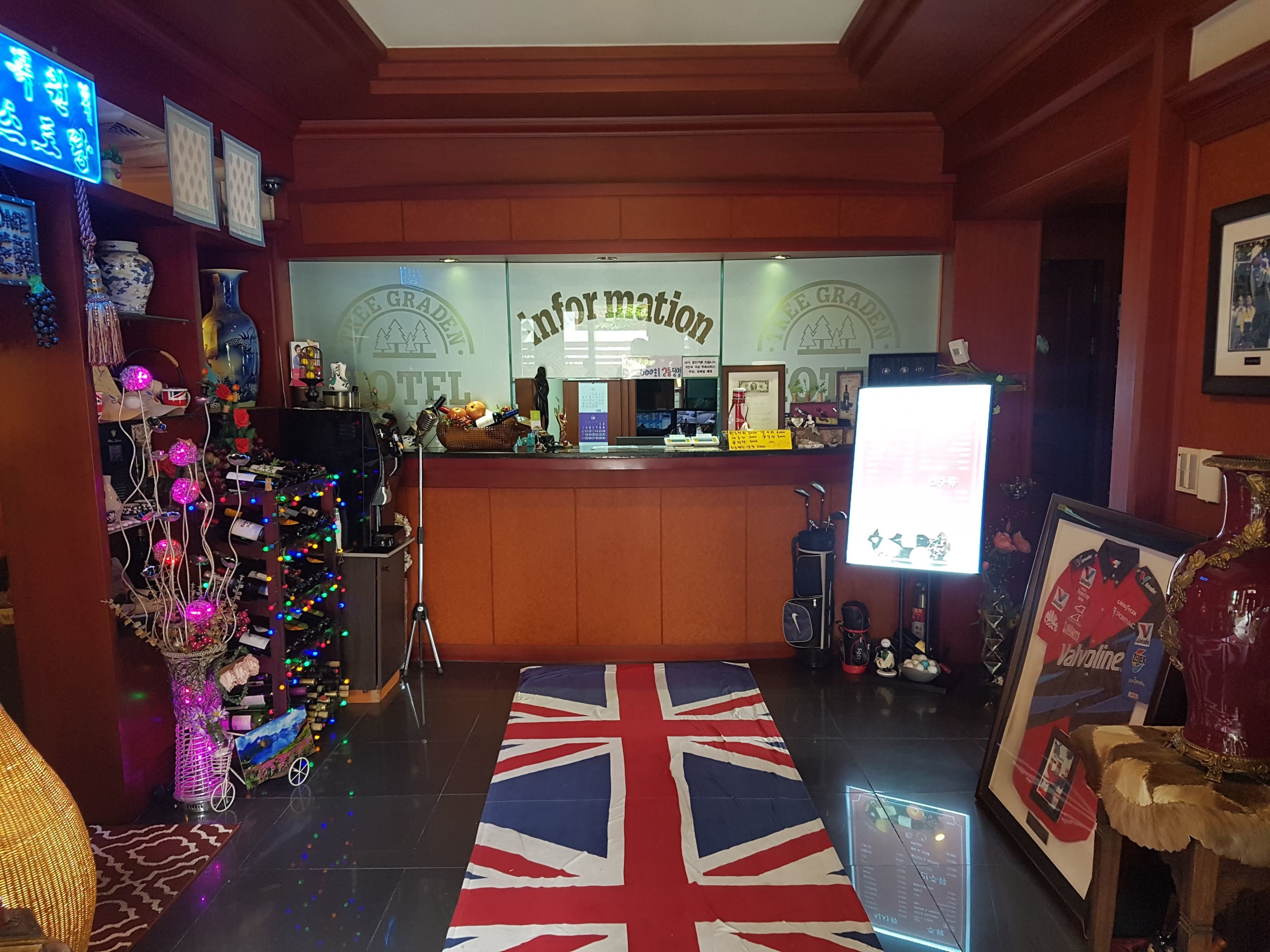 Uma área de recepção de hotel com balcão, tapete com padrão de bandeira e diversas decorações, incluindo um pinheiro de Natal e uma fotografia emoldurada.