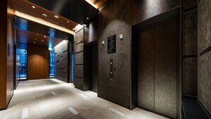 Interior moderno de elevador com parede escura, piso com piso tátil e painel de comando fixado na parede.
