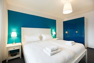 Cama bem arrumada com lençóis e toalhas brancas, mesa de cabeceira branca com abajur, e parede azul de destaque.
