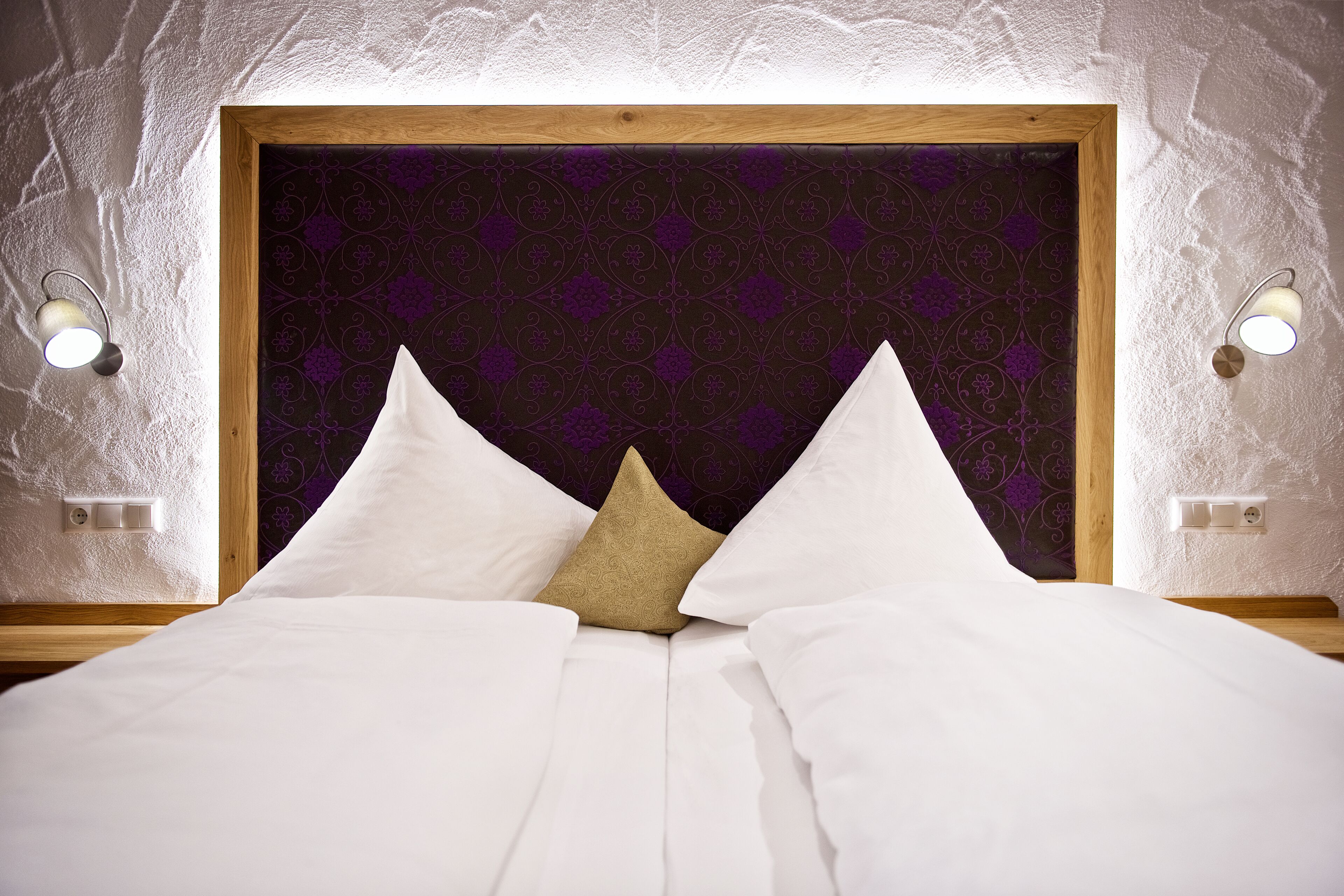 Cama bem arrumada com lençóis brancos e um único travesseiro decorativo, diante de uma parede texturizada com papel de parede estampado escuro.