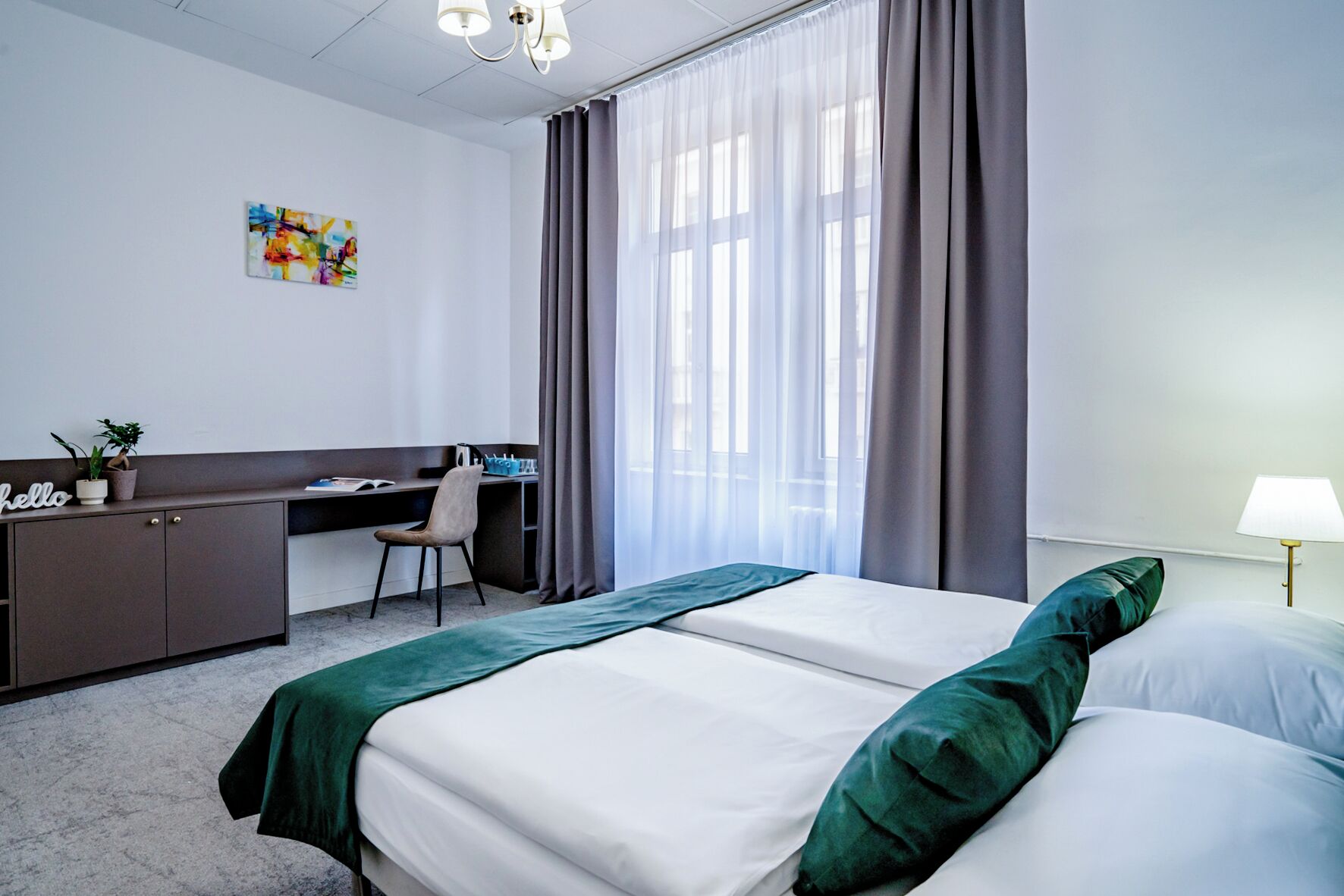 Quarto de hotel com cama, escrivaninha, cadeira, luminária e janela com cortinas.