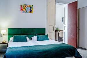 Quarto de hotel com cabeceira estofada verde, cama com roupa de cama branca e azul turquesa, luminária de cabeceira e um quadro abstrato colorido na parede.