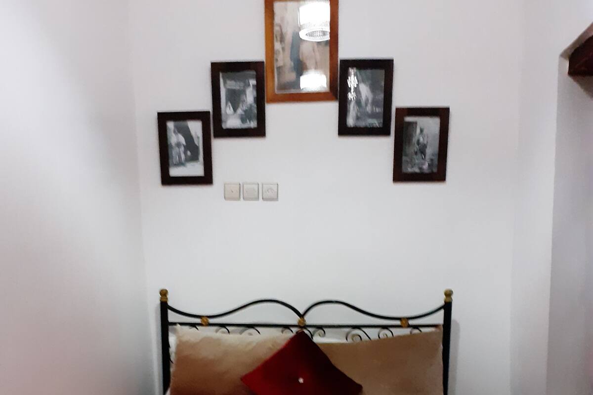 Uma cama com colcha vermelha e branca, um travesseiro vermelho e estrutura metálica preta. Há quatro quadros na parede acima da cama.